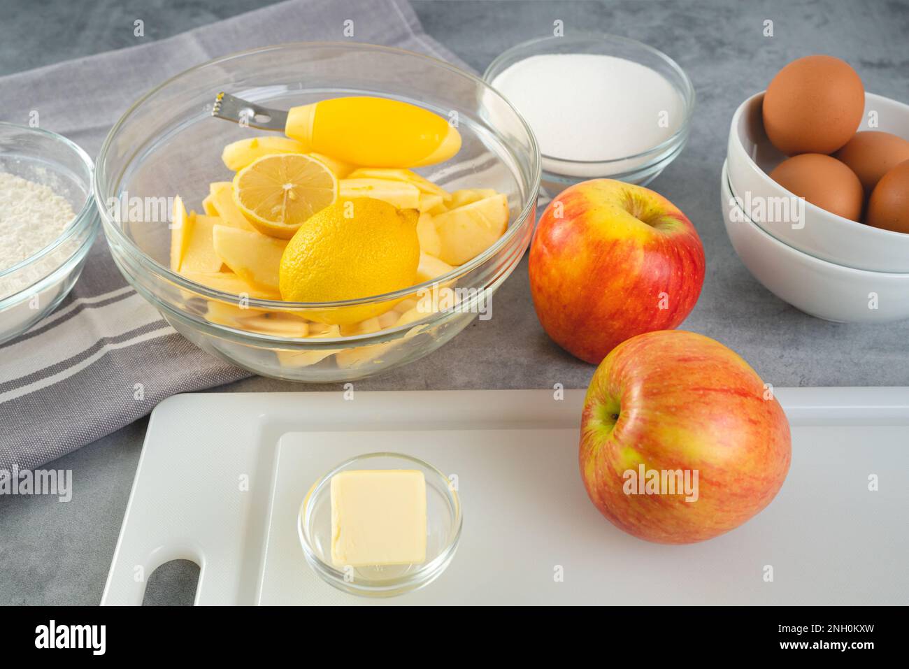 Baking pan with apple slices, lemon, eggs, sugar, and flour in bowls ...