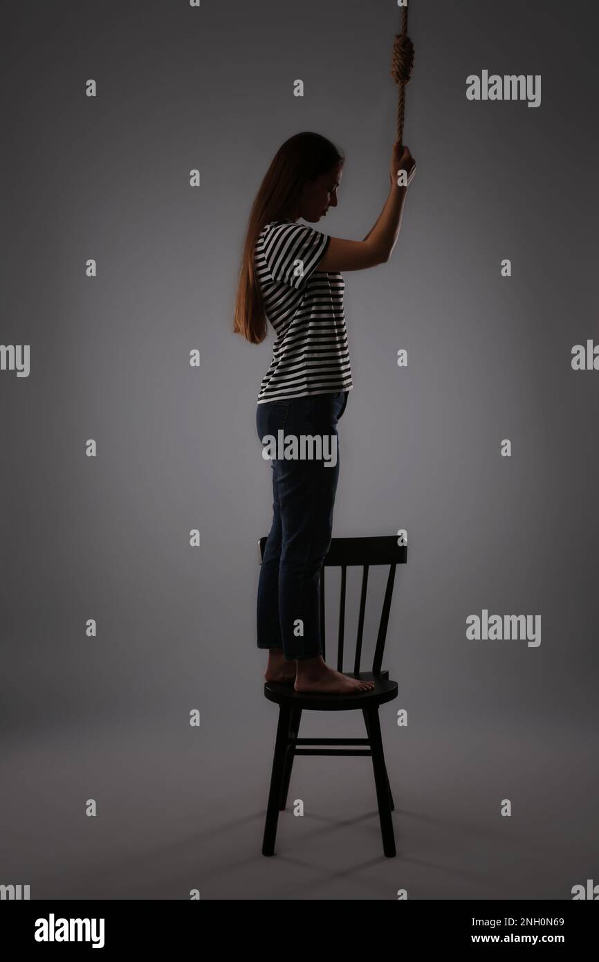 Depressed woman with rope noose standing on chair against grey ...