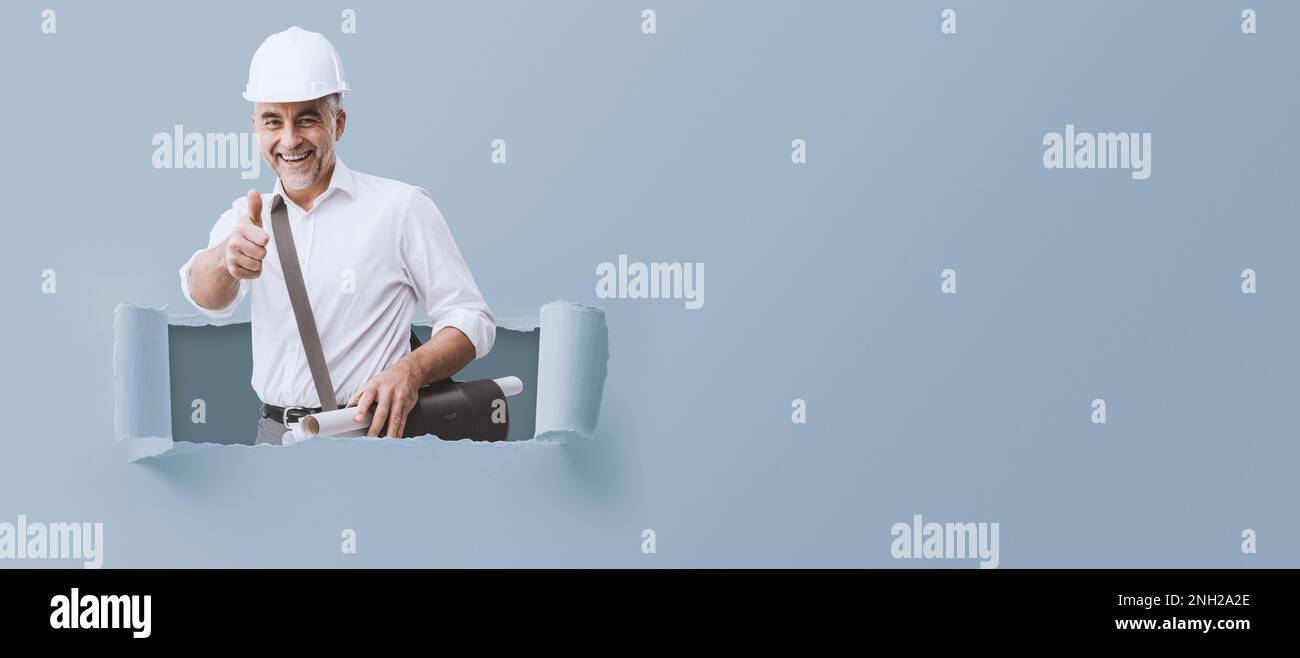 Cheerful construction engineer giving a thumbs up and carrying paper ...