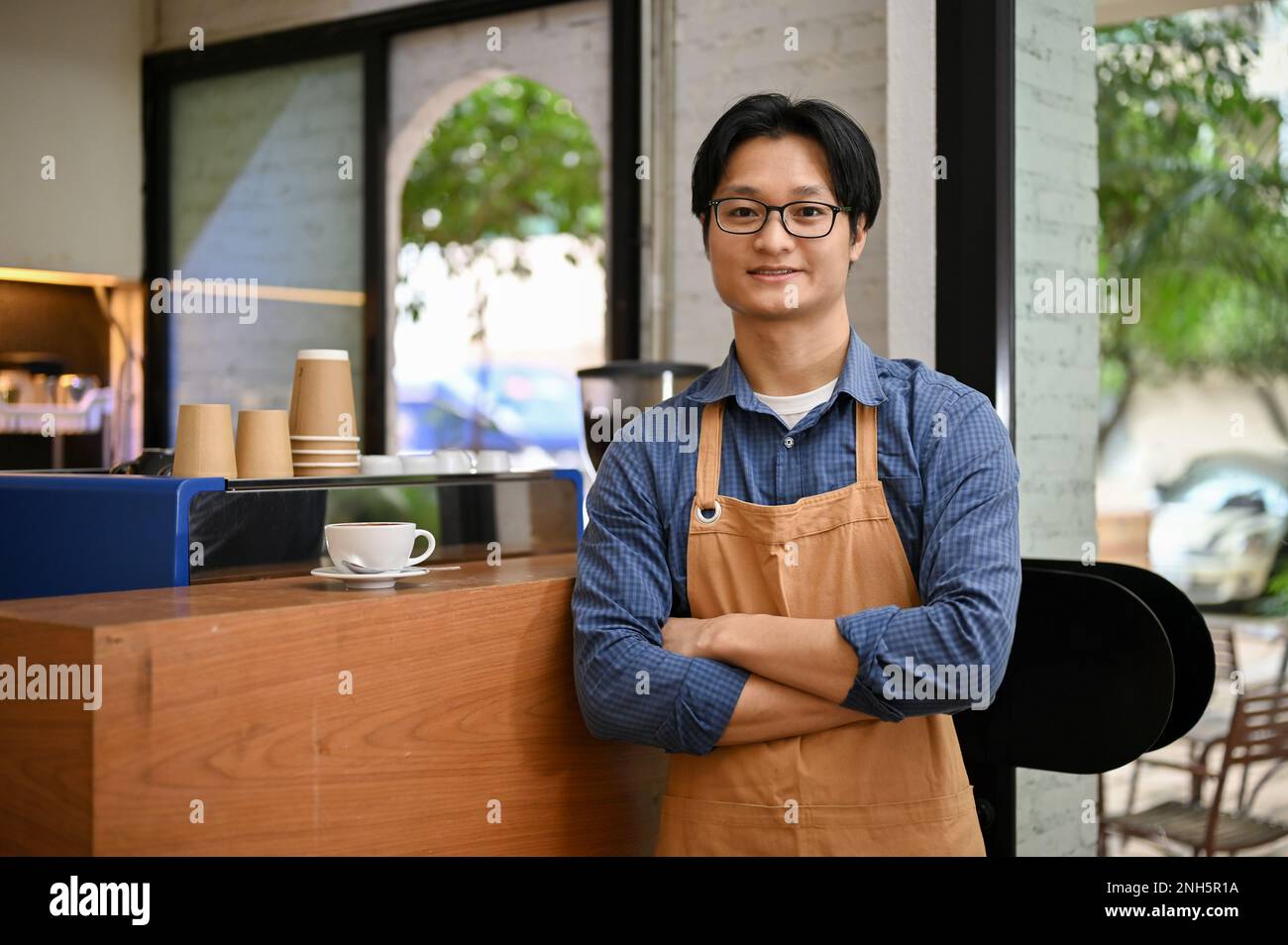 Successful and friendly Asian male barista or waiter in apron stands ...
