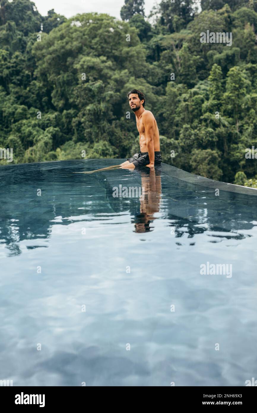 Shot of young man shirtless sitting on the edge of swimming pool at ...