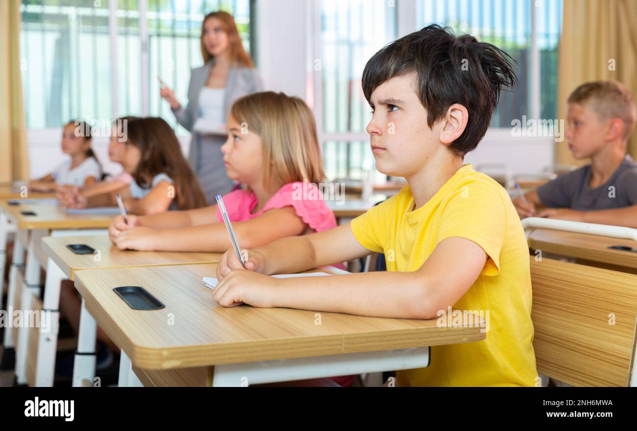 Tween boy listening to schoolteacher and writing in notebook in class ...
