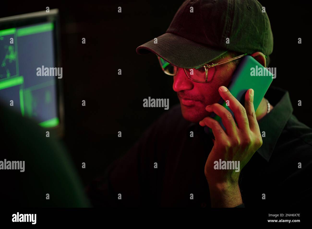 Hacker in cap and glasses talking on phone, when working in dark room ...