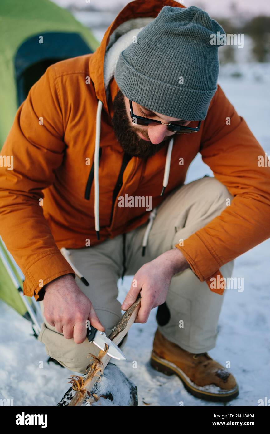 A man prepares wood wool with a knife for starting a fire in a winter ...