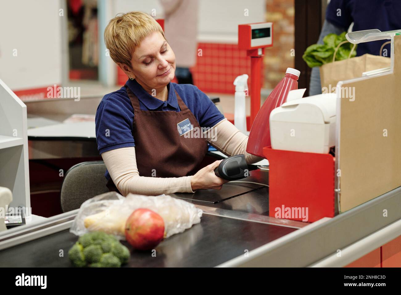 Blond pretty female cashier in uniform scanning price of product in red ...