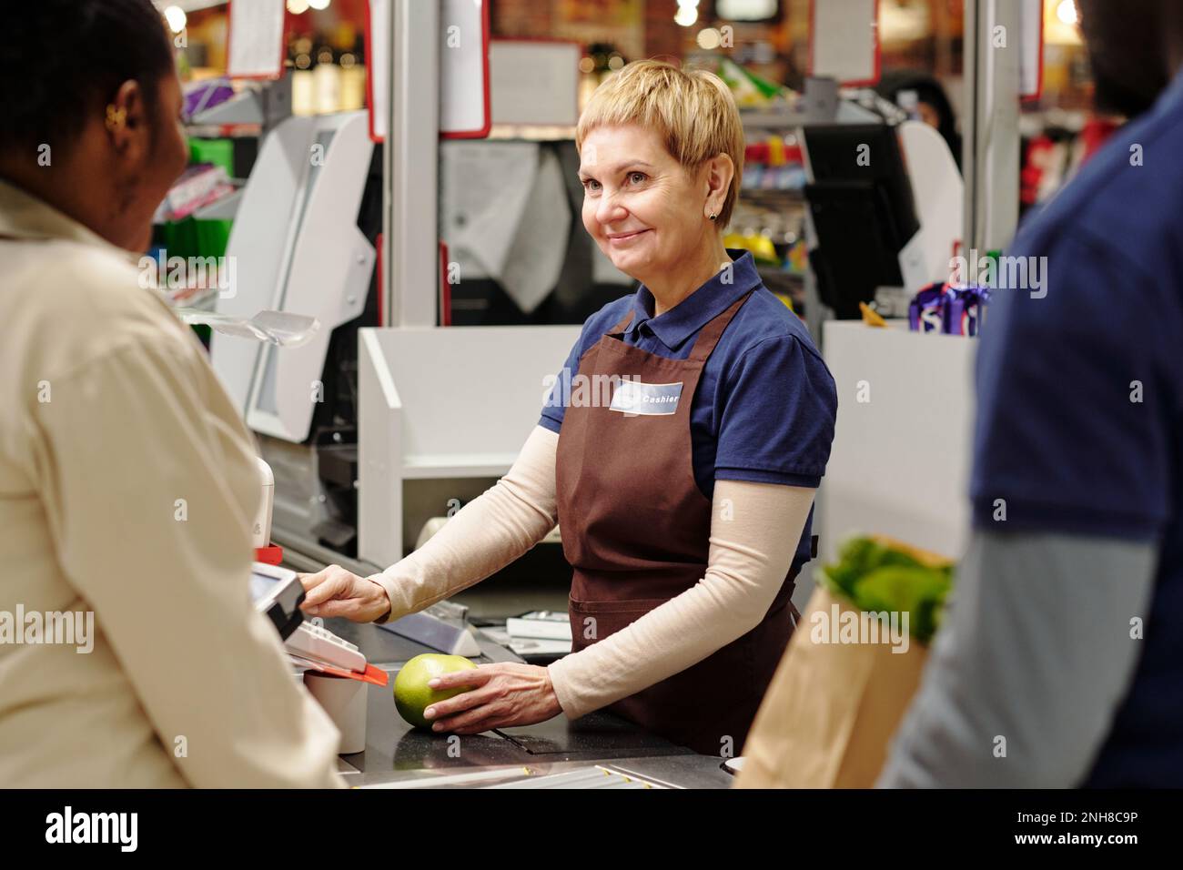 Happy blond mature cashier looking at African American female consumer ...