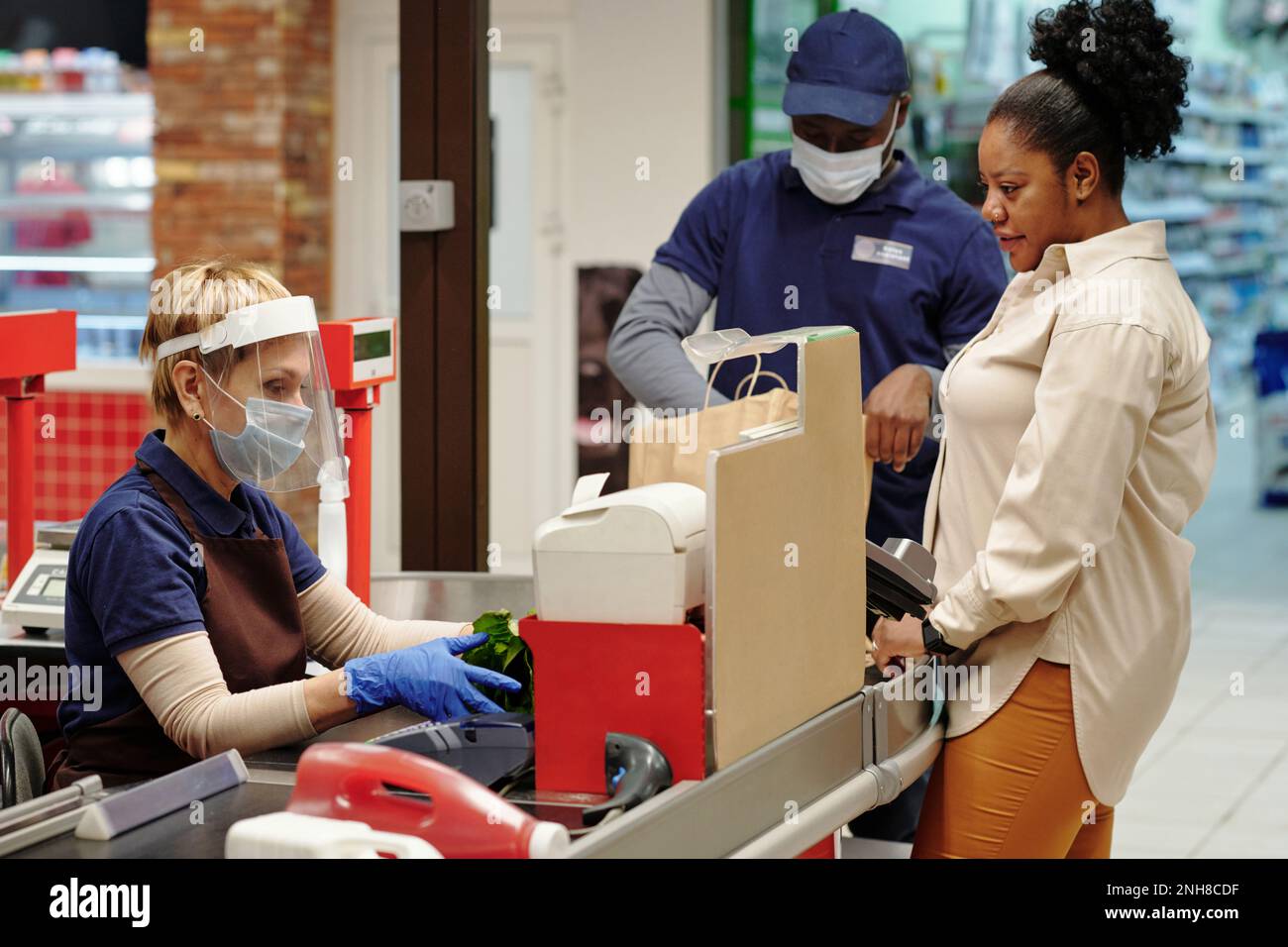 Mature female cashier in protective mask and screen scanning food ...