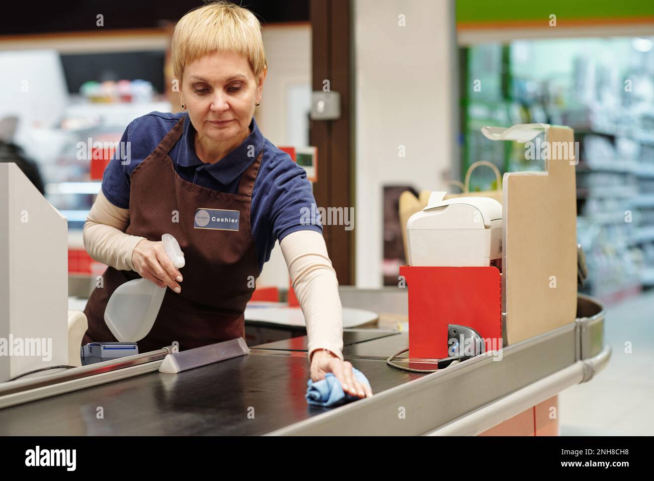Mature female cashier in uniform spraying sanitizer on checkout line ...