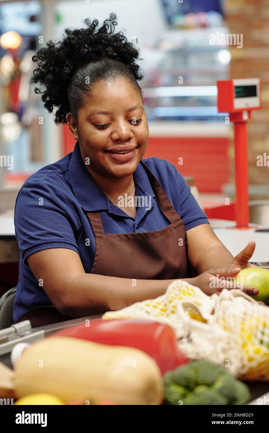 Happy young female cashier in uniform sitting by checkout counter in ...