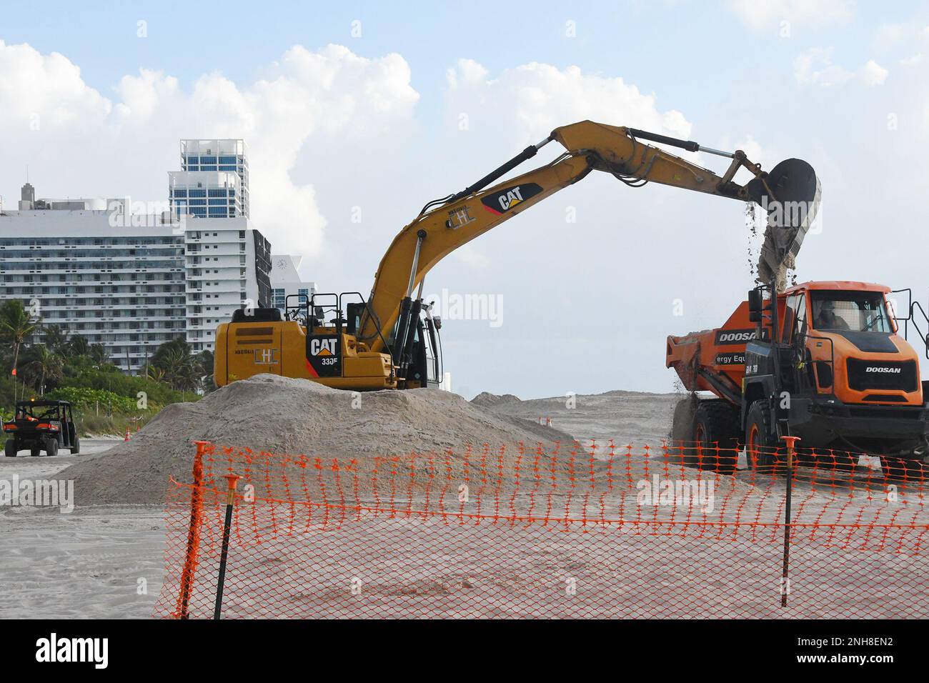 An excavator sits on top a large pile of beach sand and loads sand onto a dump truck at Indian ...