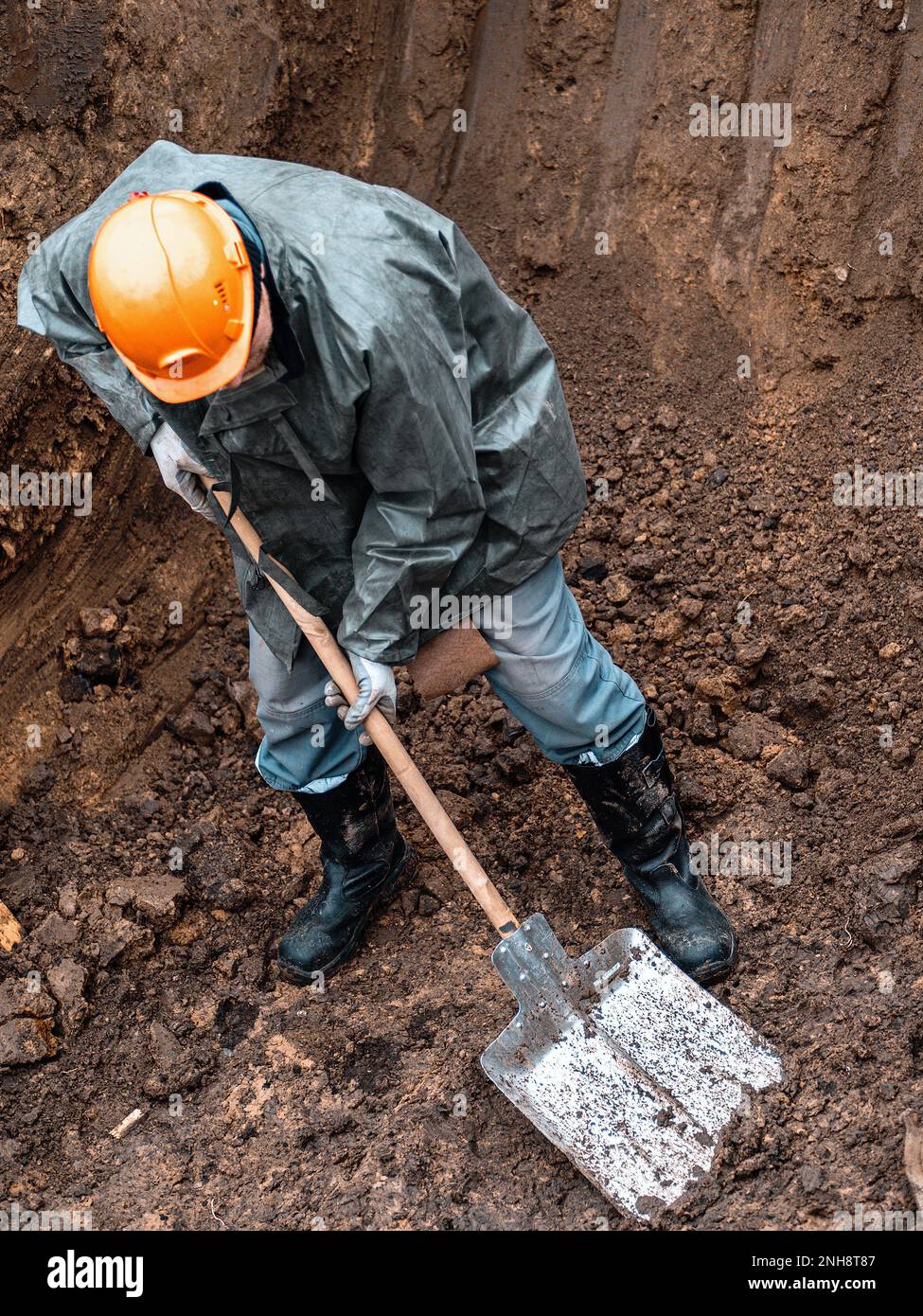 Man in helmet, raincoat and work clothes with shovel in pit digs hole ...