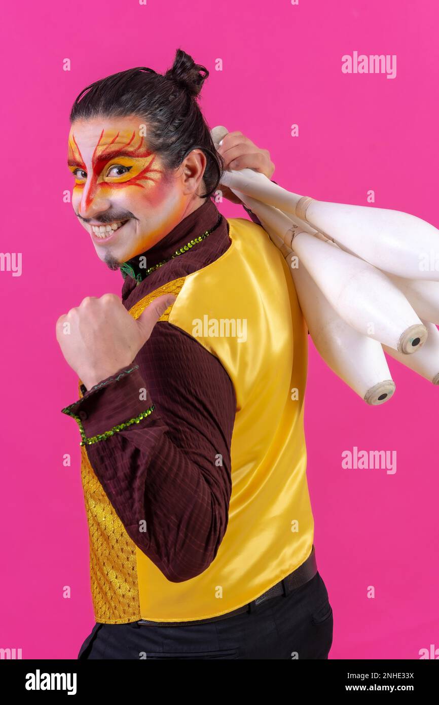 Juggler in a vest and with painted face juggling with maces on a pink ...
