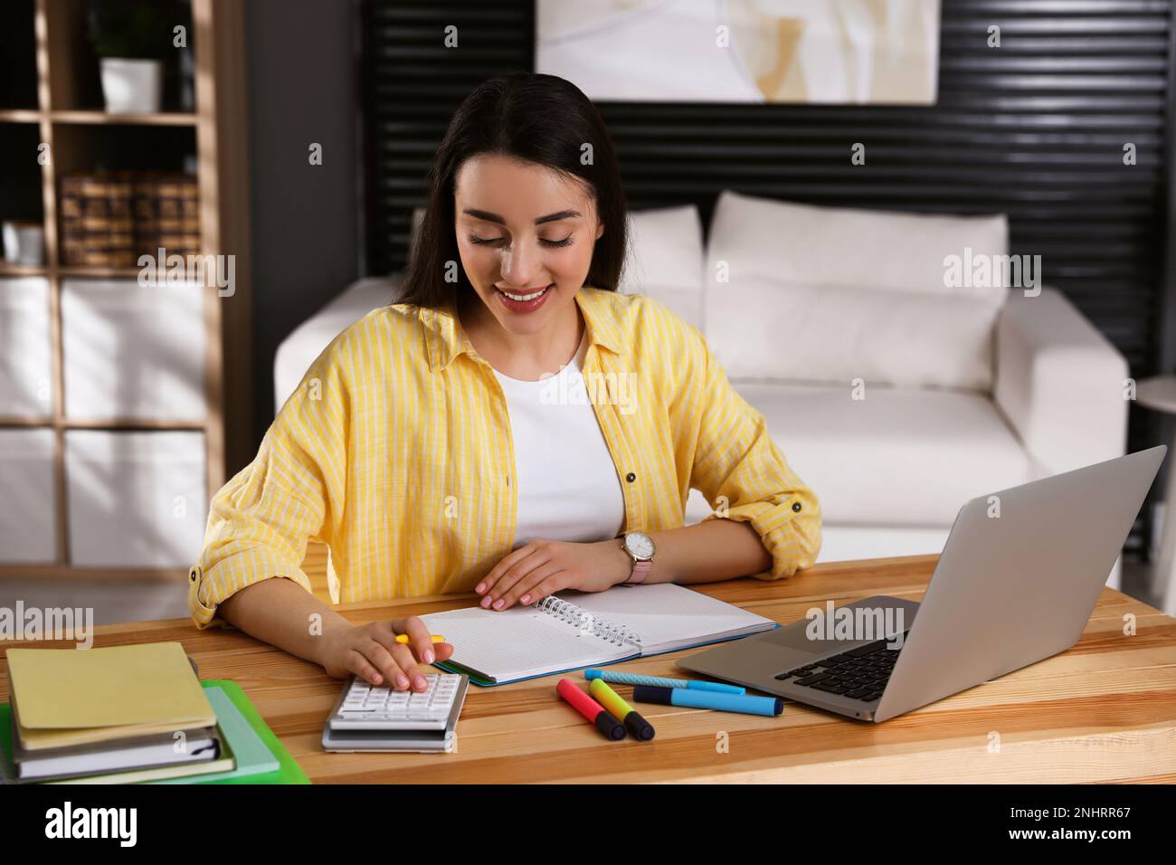 Young woman counting on calculator during webinar at table in room ...