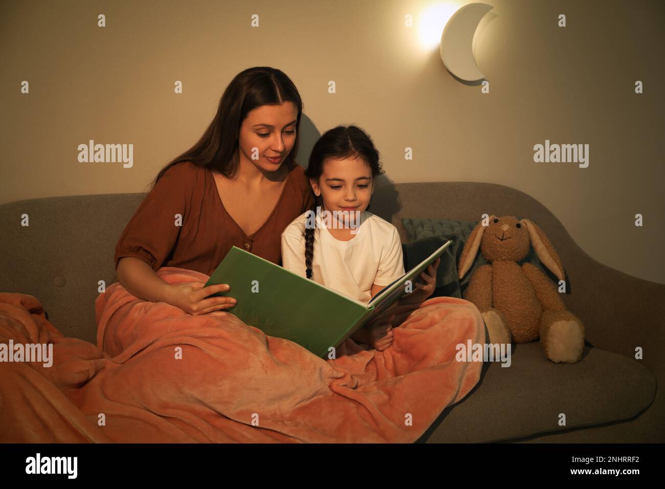 Little girl with mother reading book in living room lit by night lamp ...
