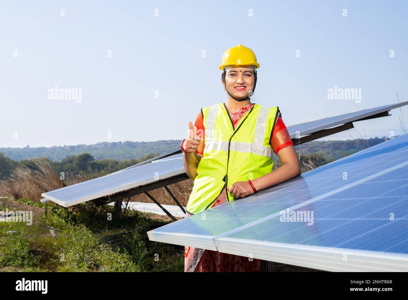 Portrait of Young indian woman technician wearing yellow hard hat standing near solar panels ...
