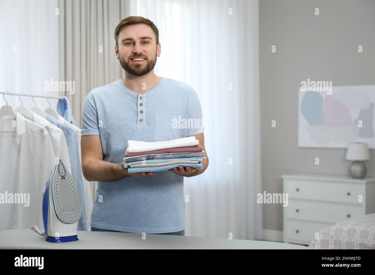 Man with folded clothes after ironing near board at home. Space for ...