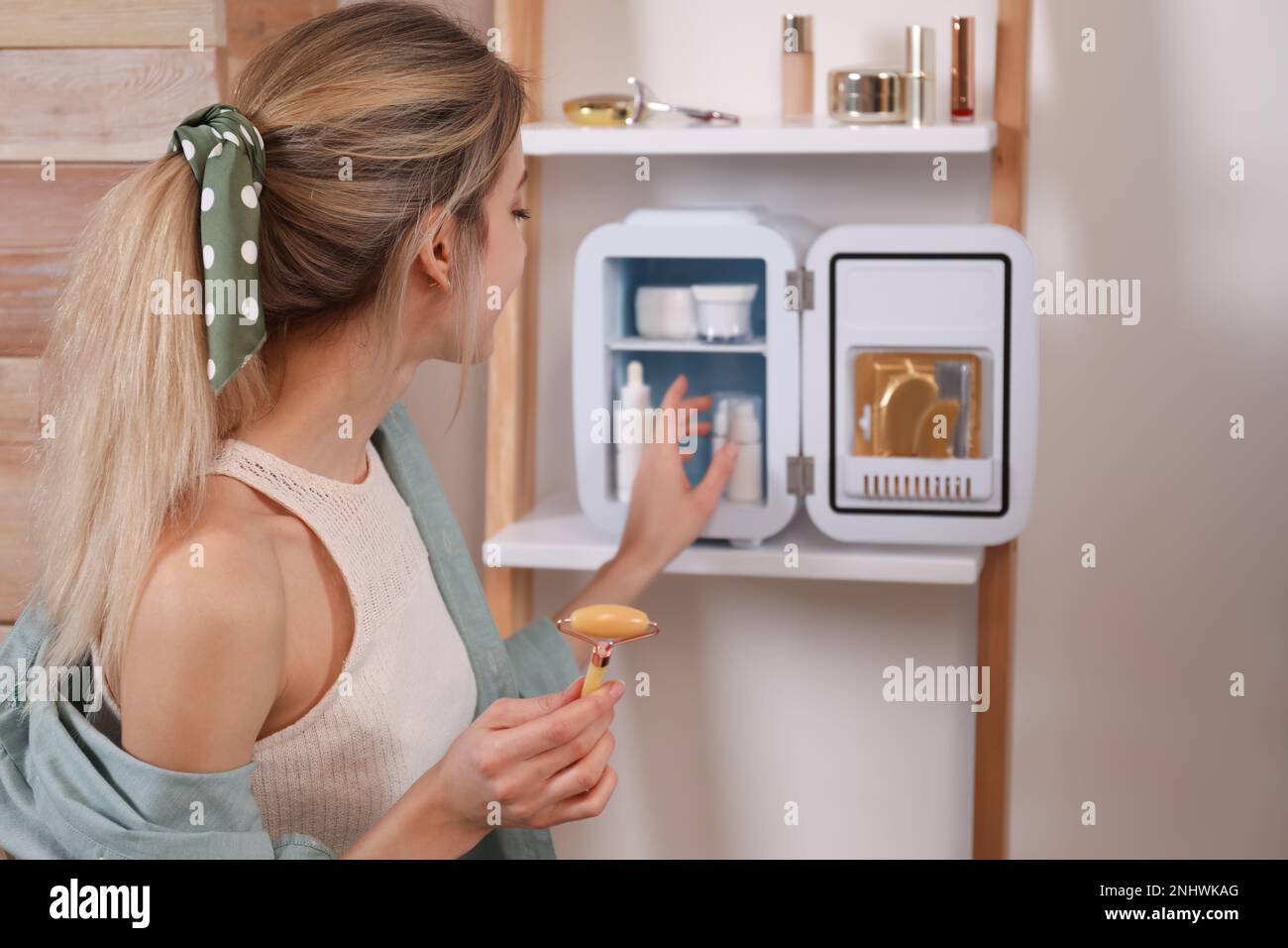 Woman with face roller taking cosmetic product from mini fridge indoors ...