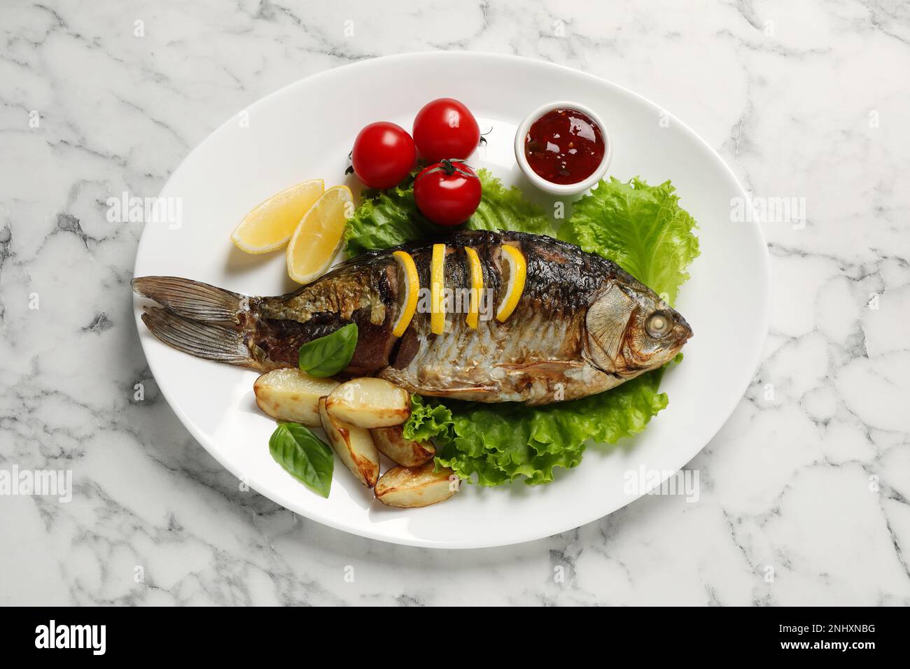 Tasty homemade roasted crucian carp with garnish on white marble table ...