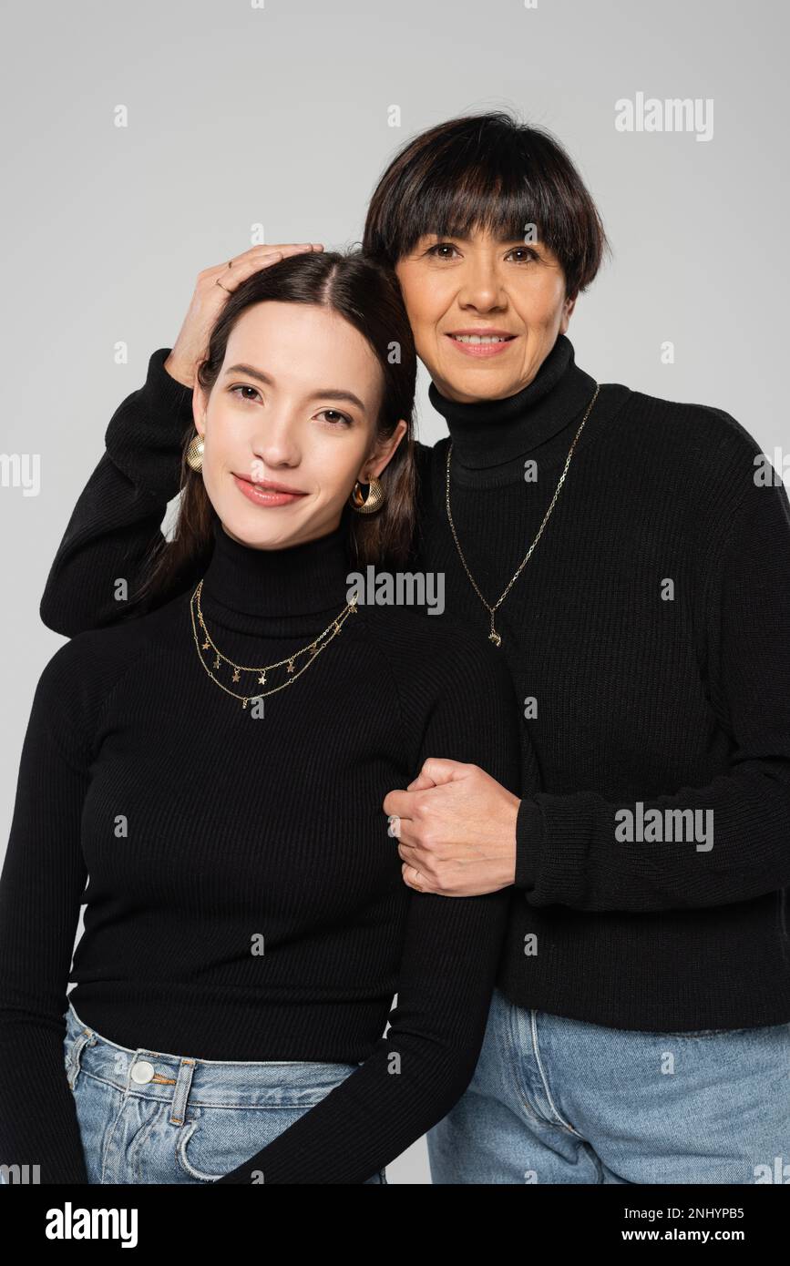 cheerful asian woman hugging young daughter wearing black turtleneck and necklaces isolated on ...
