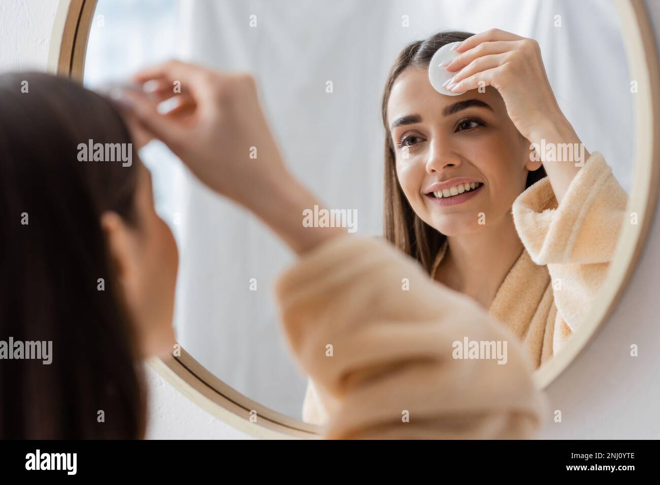 reflection of smiling young woman cleansing face with cotton pad in ...