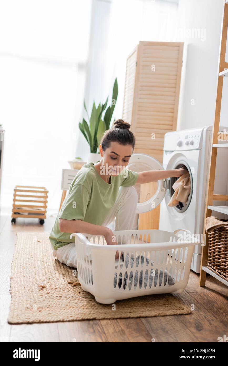 Brunette woman putting clothes in washing machine near basket in ...