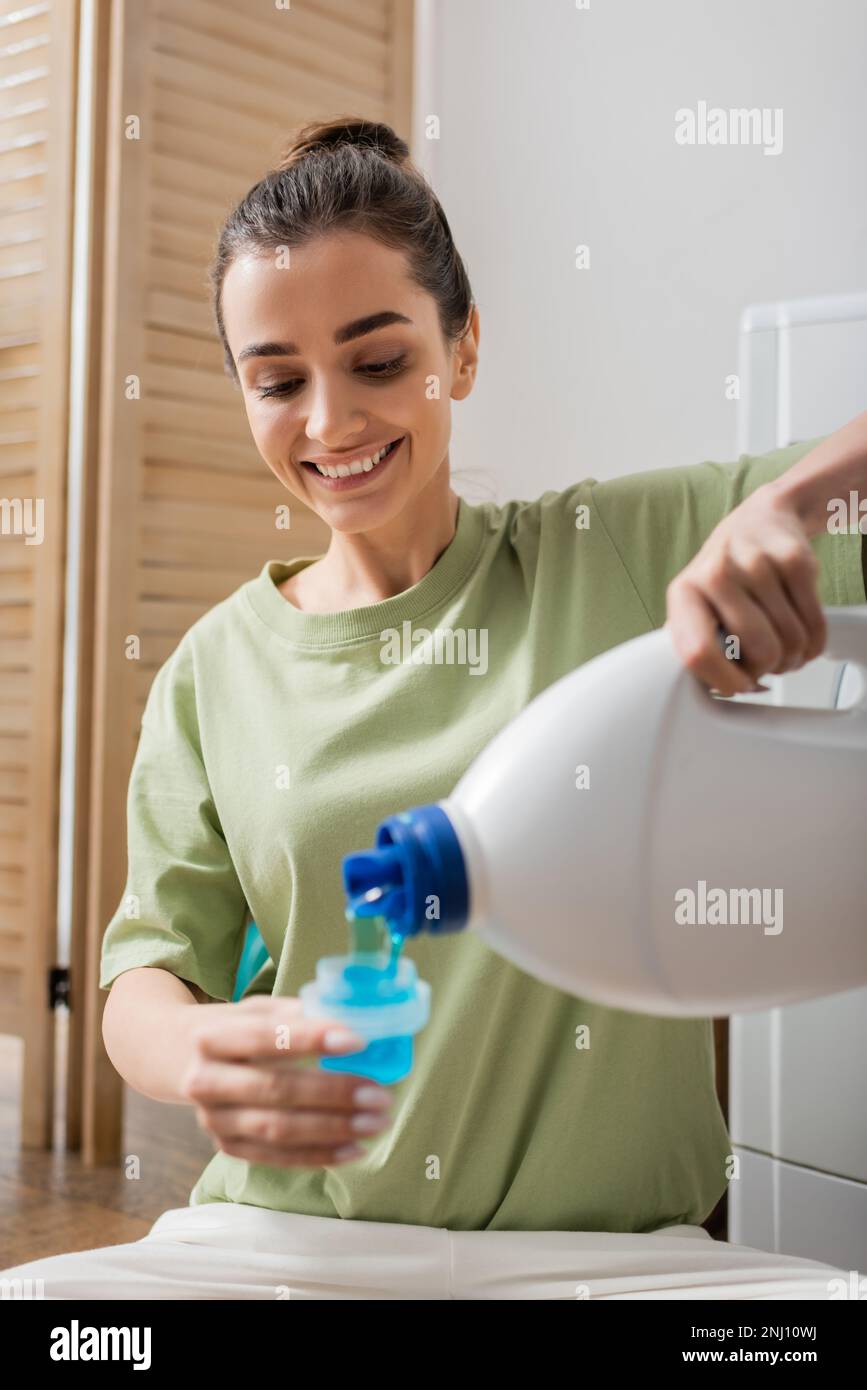 Cheerful brunette woman pouring blue washing liquid in laundry room ...