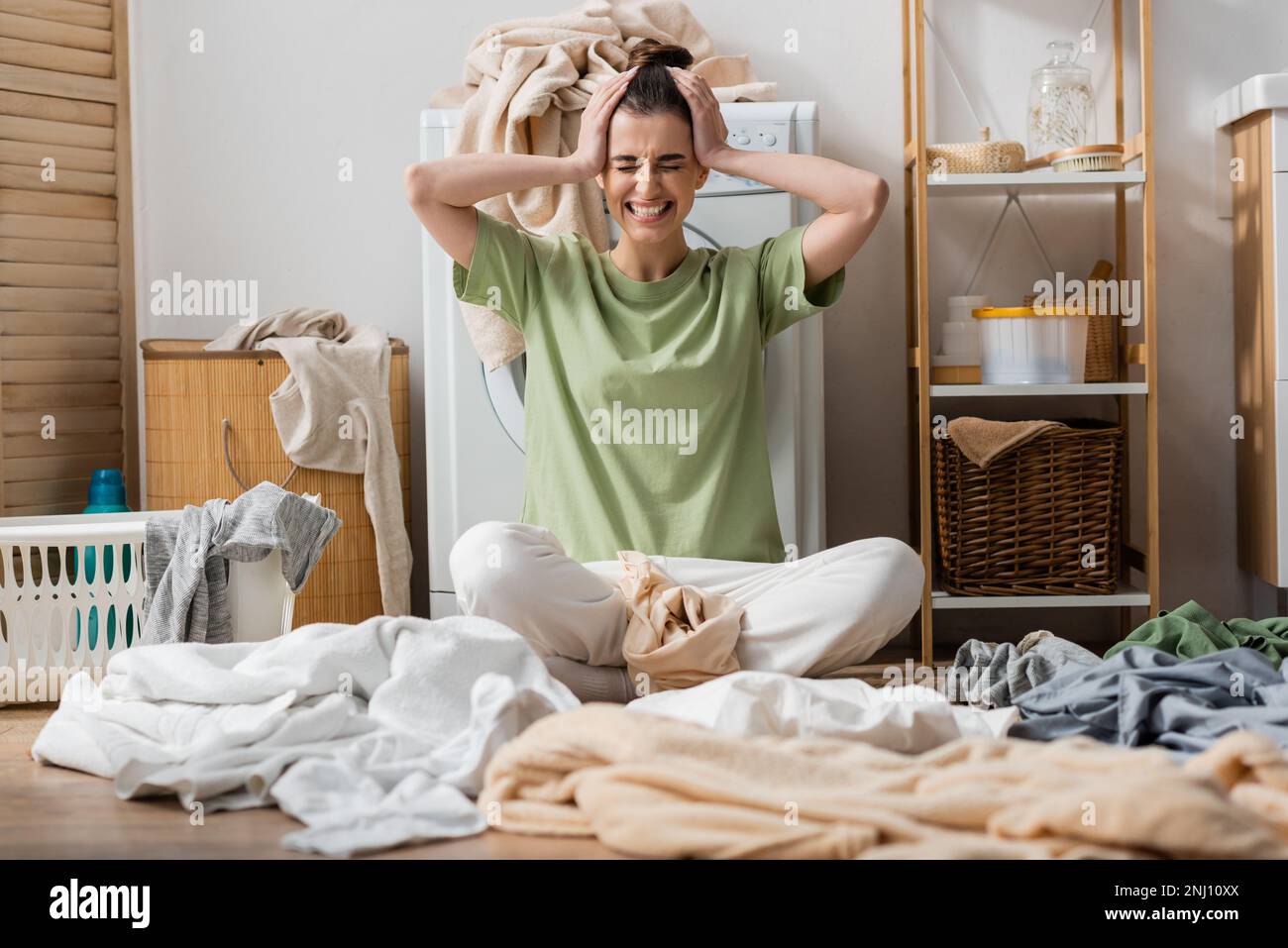 stressed young woman sitting near clothes on floor in laundry room ...