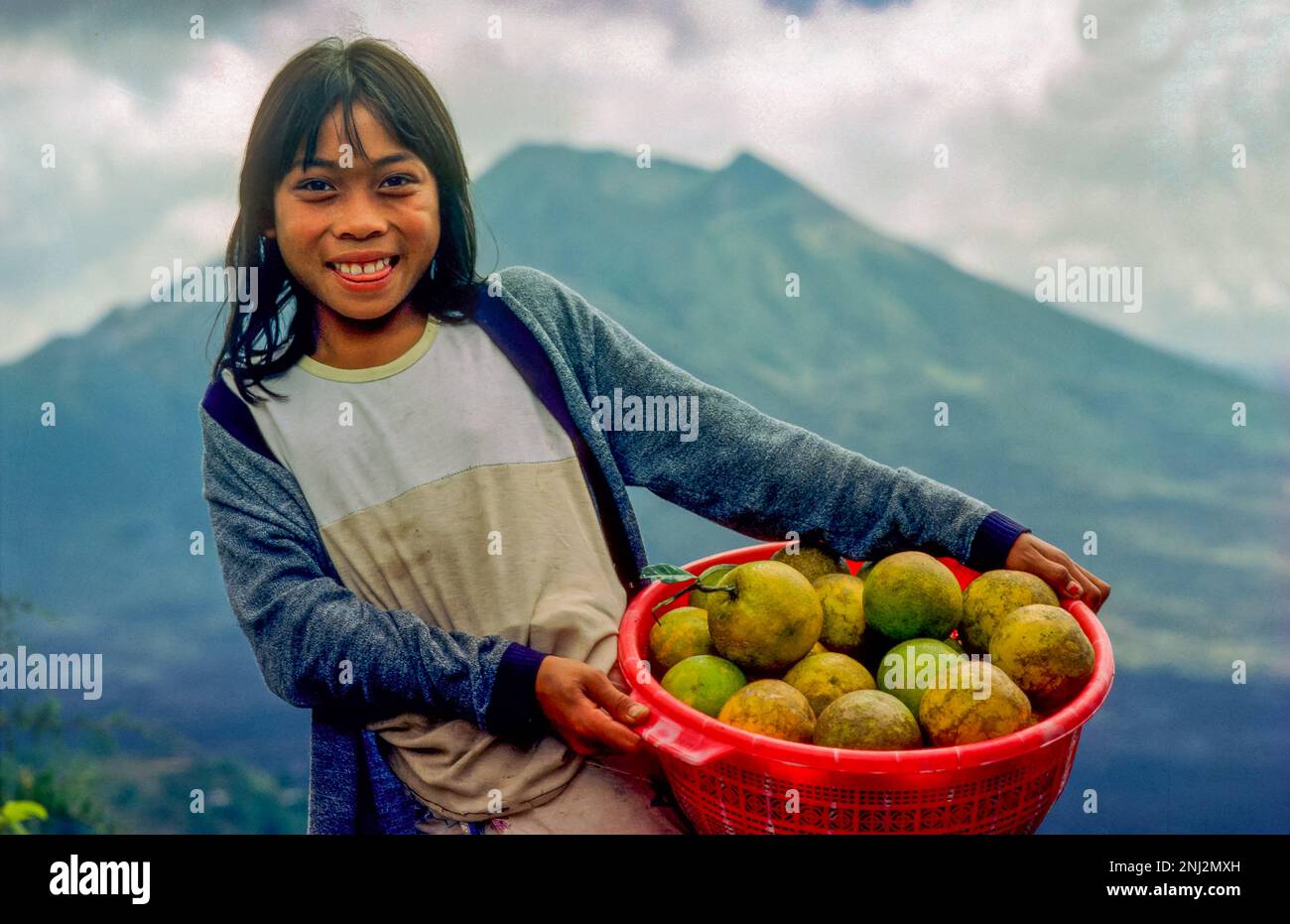 Indonesia, Bali. Girl carrying oranges in front of volcano Stock Photo ...