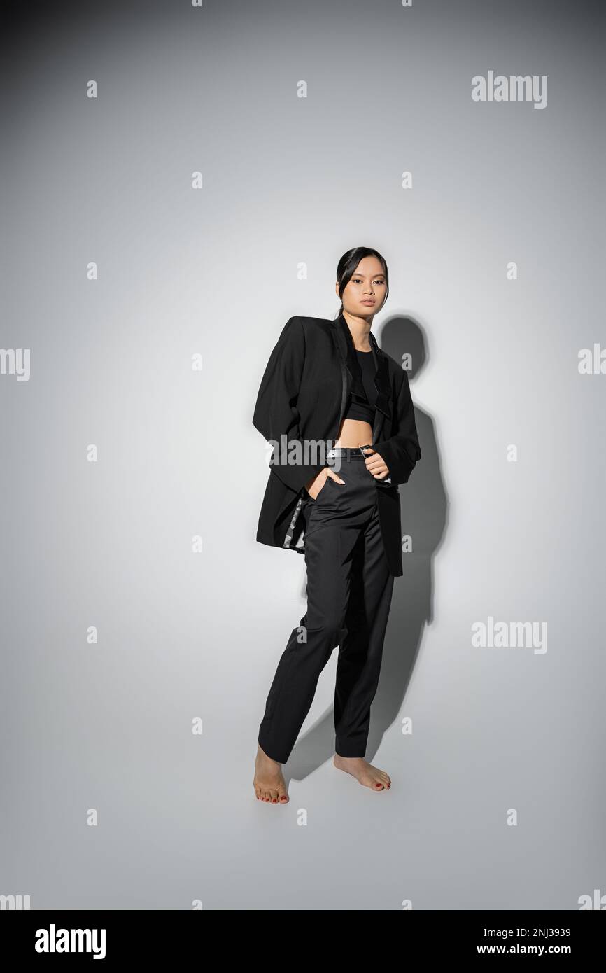 full length of barefoot asian woman in black pantsuit posing with hand ...