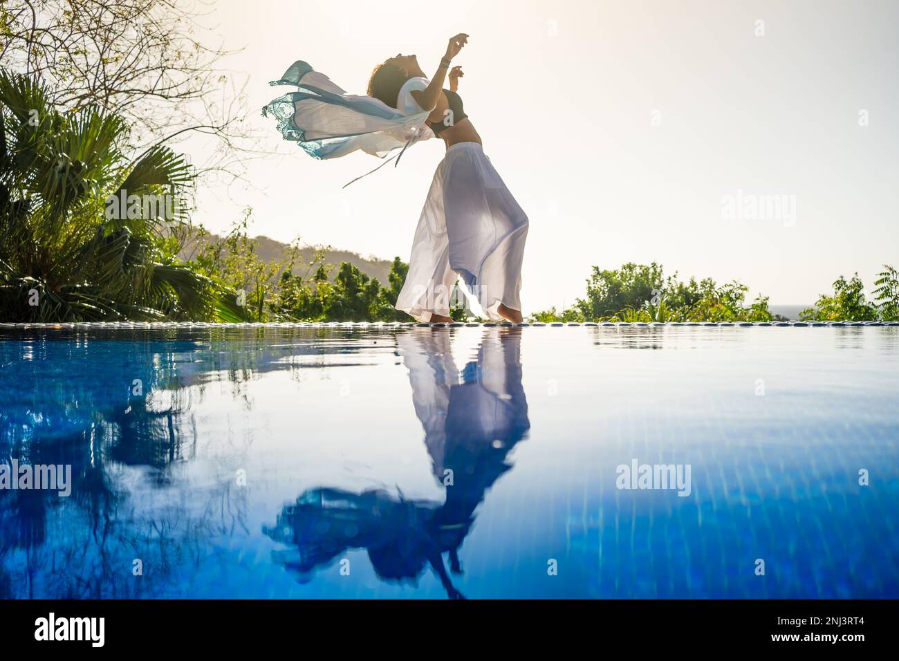 Woman dancing next to an outdoor tropical pool with the image reflected ...