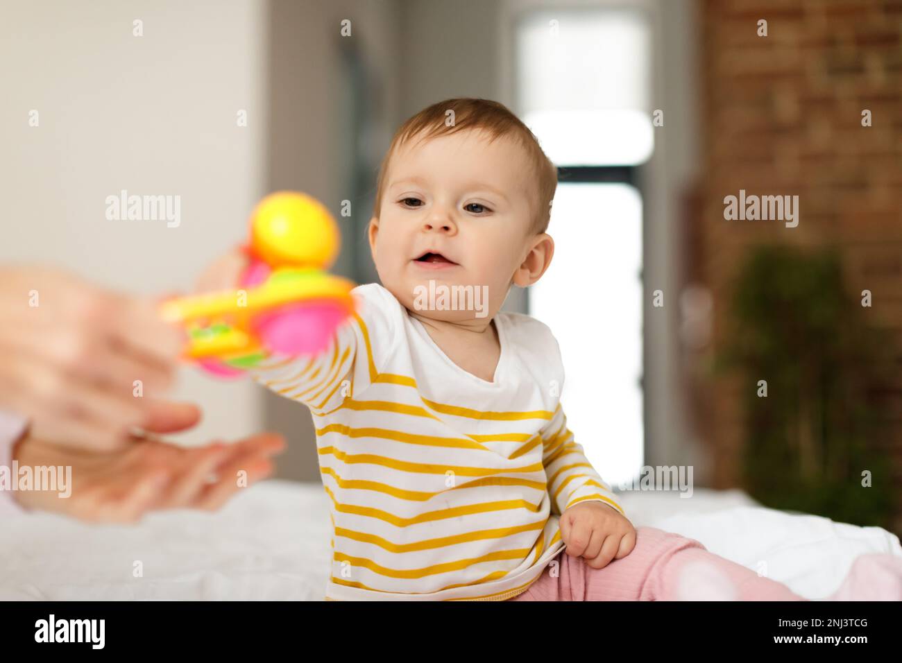 Cute baby girl taking rattle toy from mother hands, sitting on bed ...