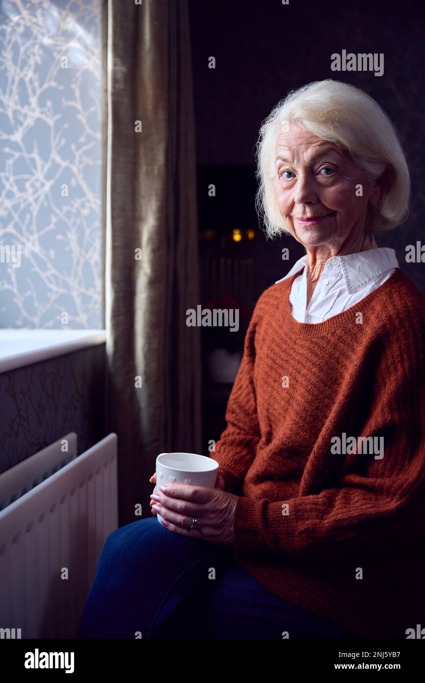 Senior Woman With Hot Drink Trying To Keep Warm By Radiator At Home In ...