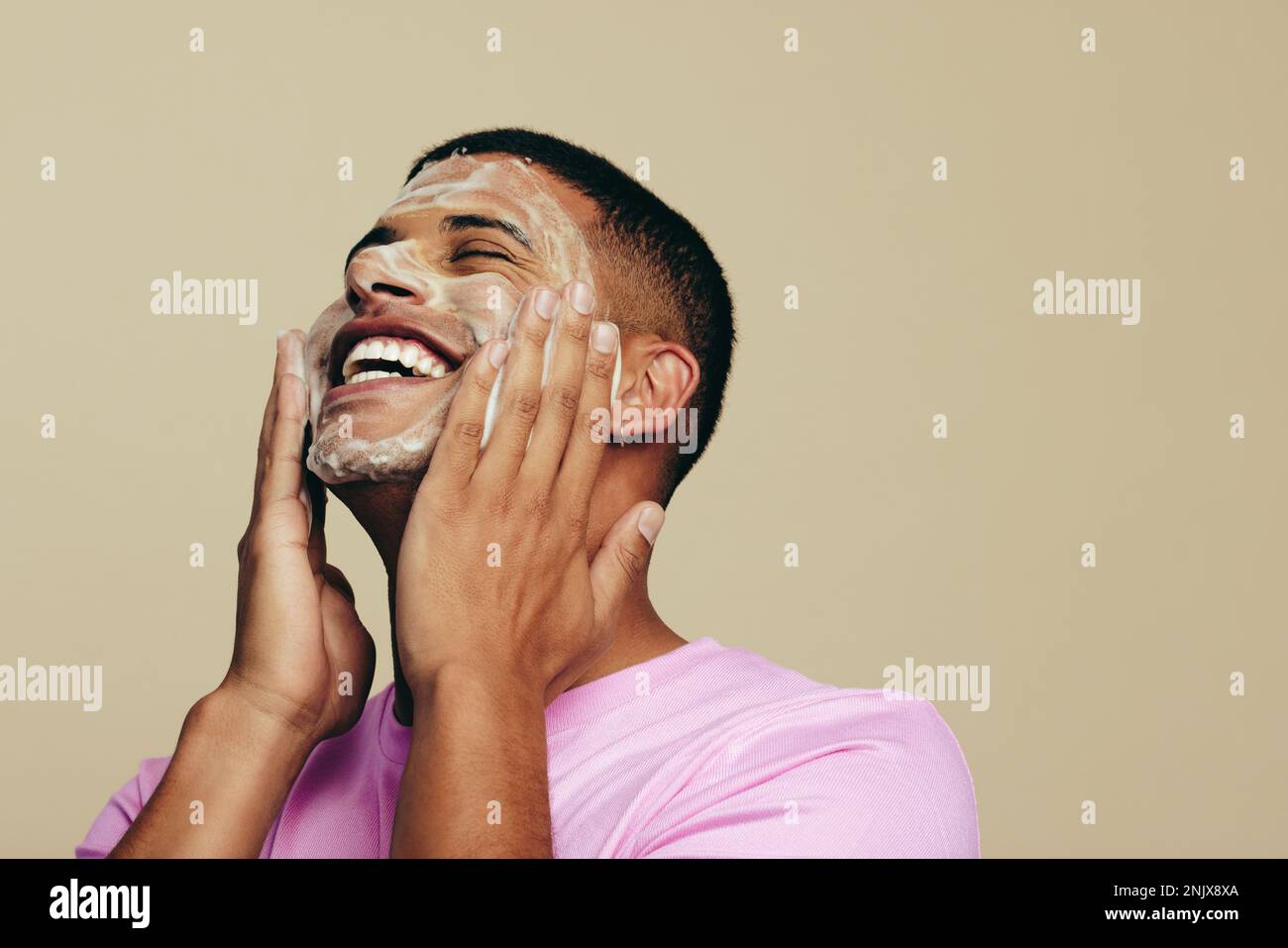Man smiling happily as he applies a face wash product in a studio, gently massaging his skin ...