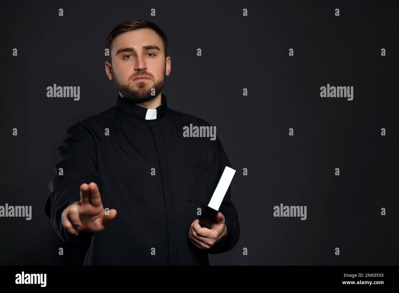 Priest with Bible making blessing gesture on black background. Space ...