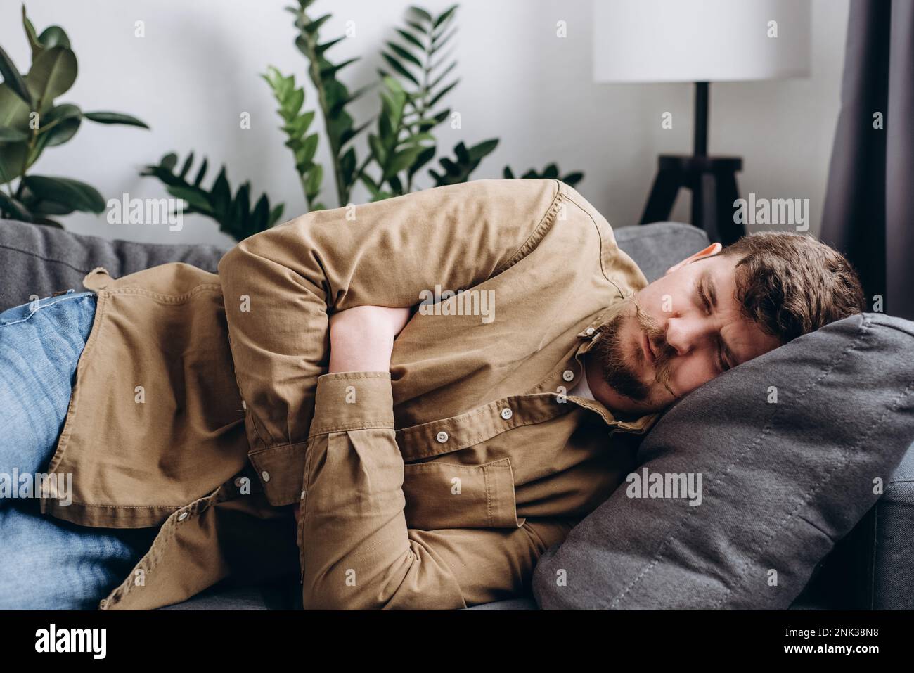 Sad young man lying on grey couch at home, suddenly feeling strong ...