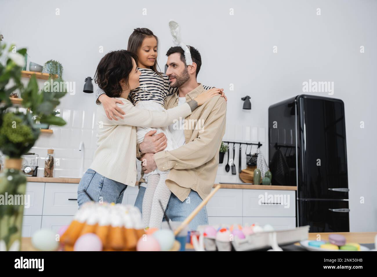 Positive parents hugging daughter near blurred Easter eggs and cake in ...
