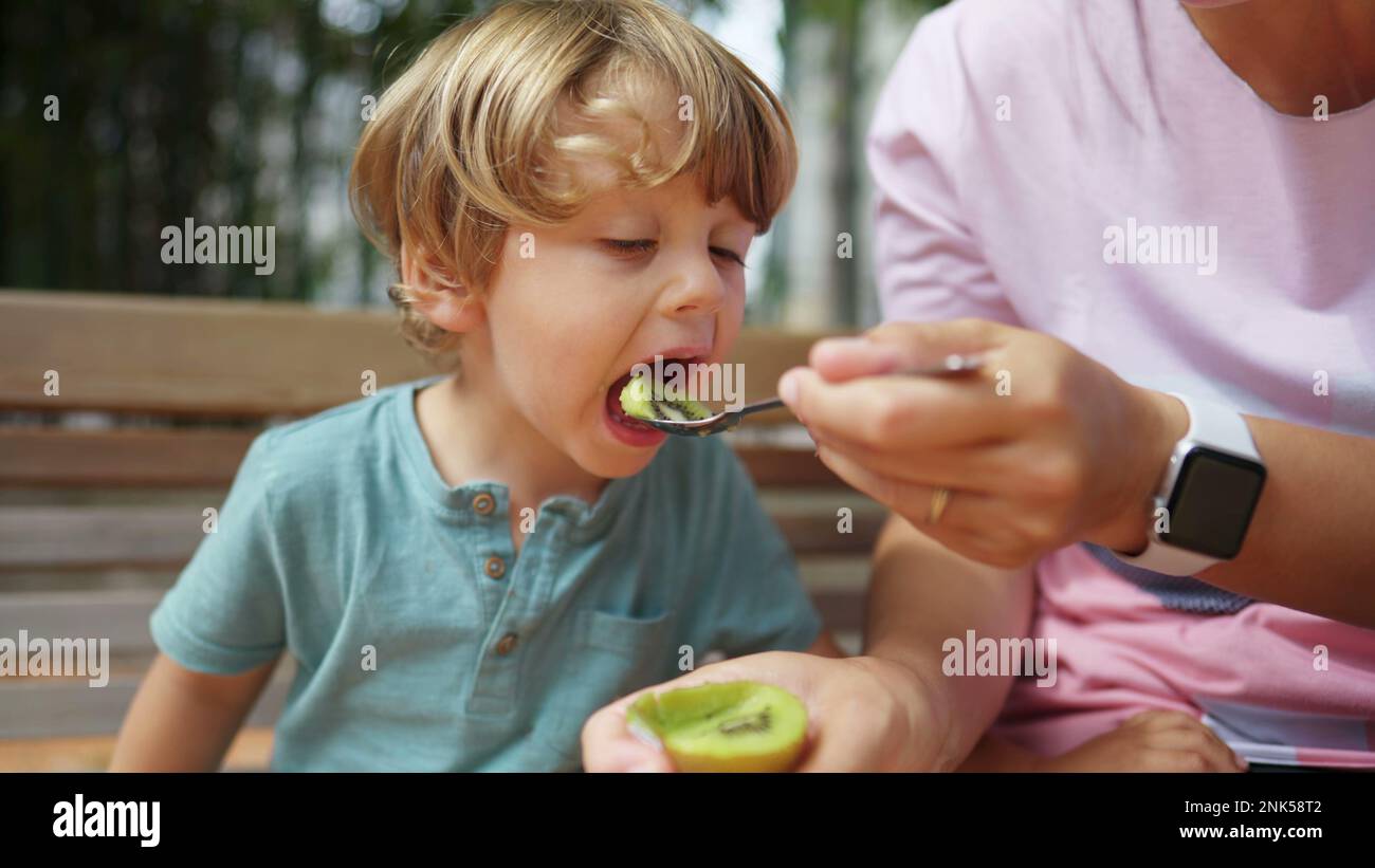 Child snacking kiwi fruit outside sitting on park bench. Closeup mother ...