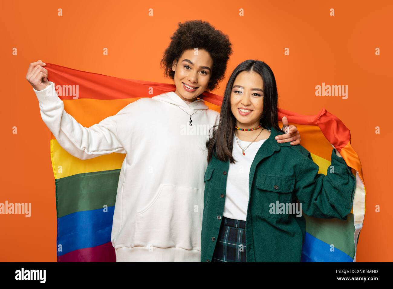 positive and trendy asian and african american women holding lgbt flag isolated on orange,stock ...