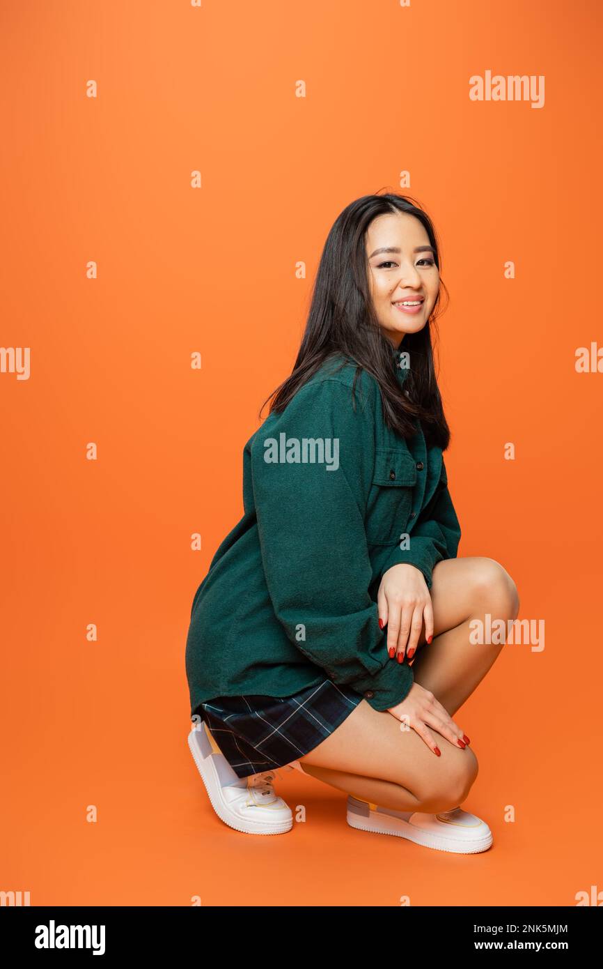 full length of happy asian woman in trendy outfit posing on haunches on ...