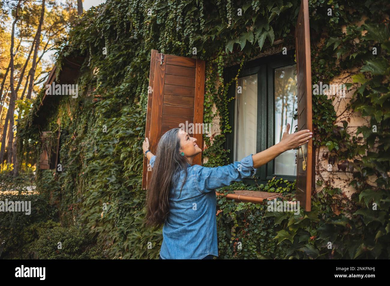 Mature woman closing window shutters of house with overgrown facade ...