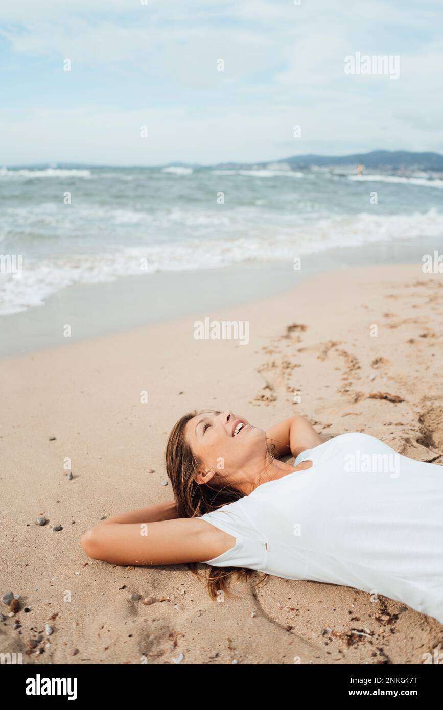 Woman with hands behind head lying on sand by sea at beach Stock Photo ...