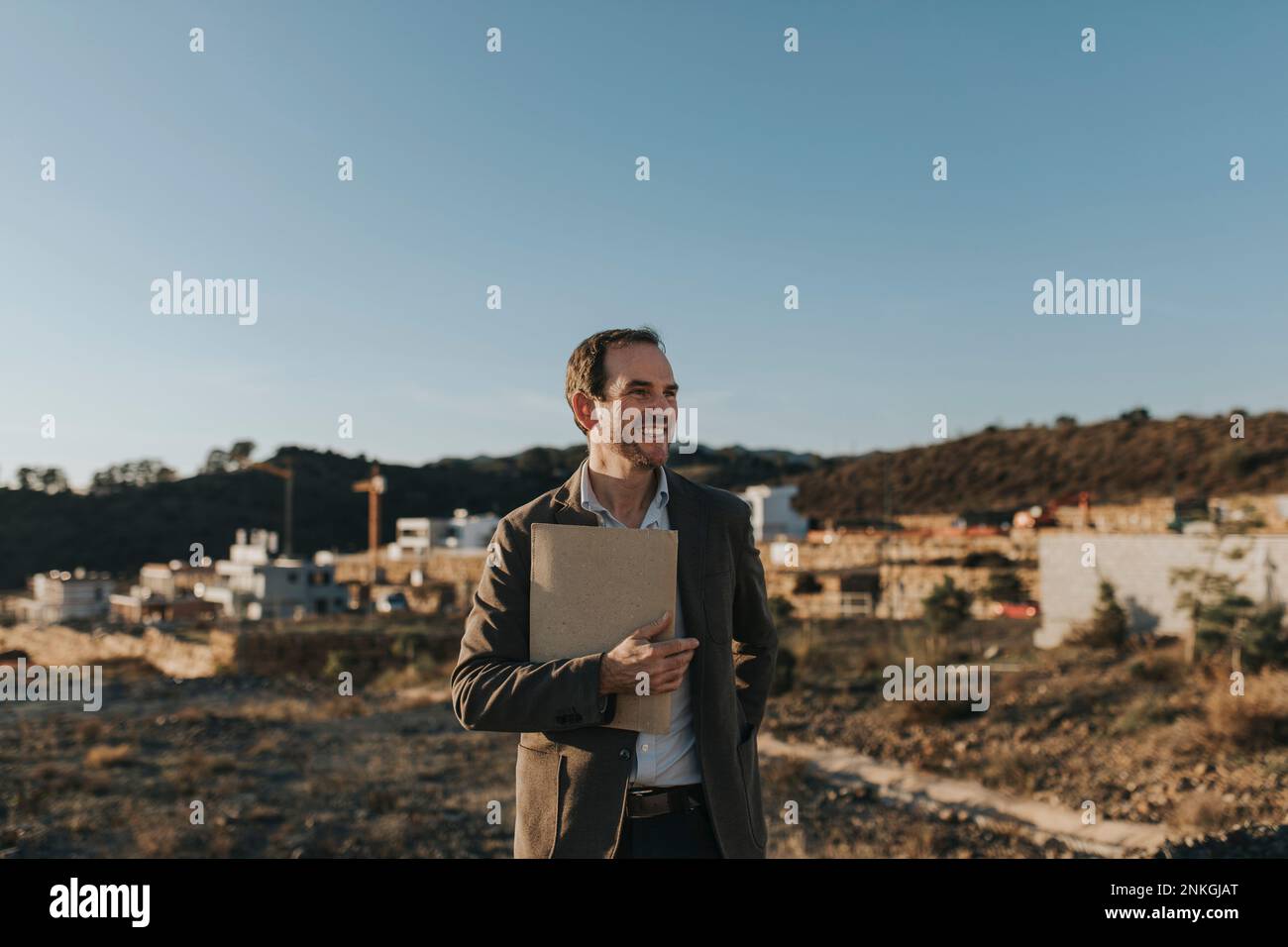Smiling man holding file standing at empty area for residential ...
