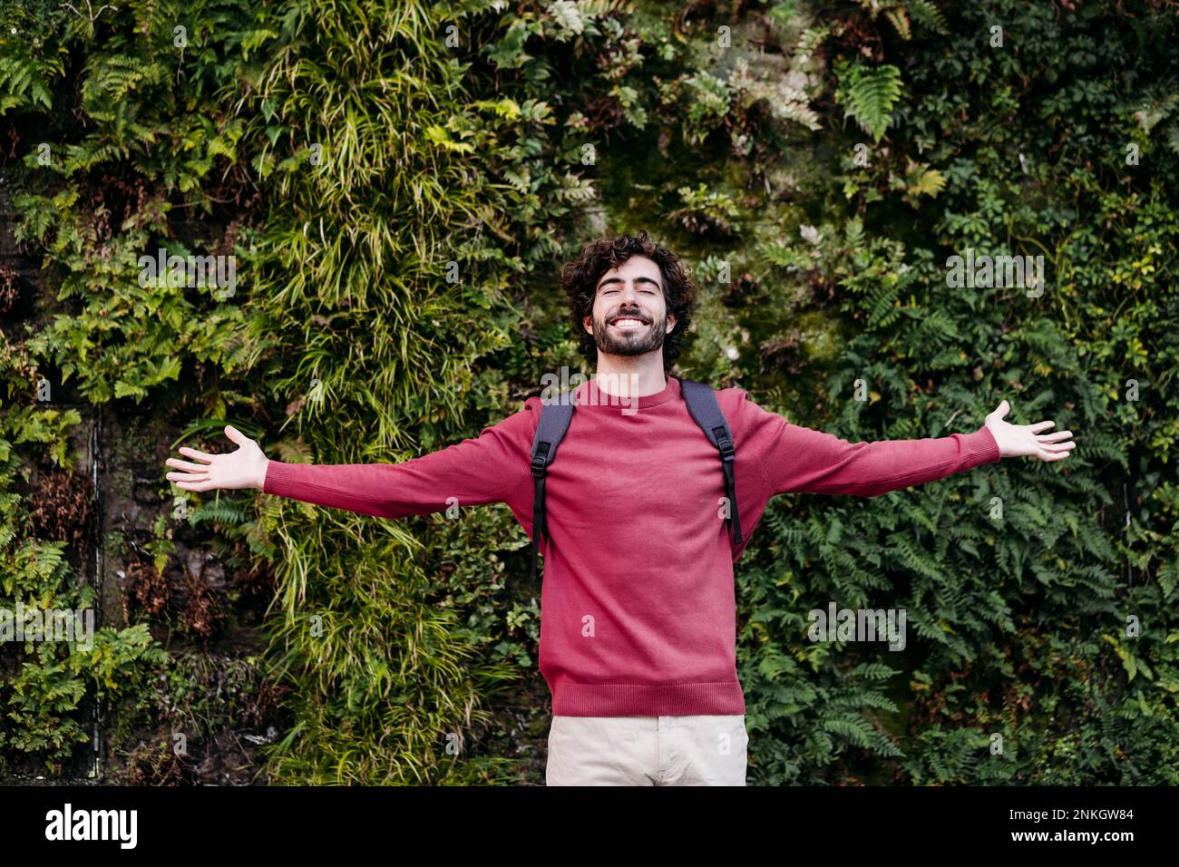 Carefree young man with arms outstretched standing in front of plants ...