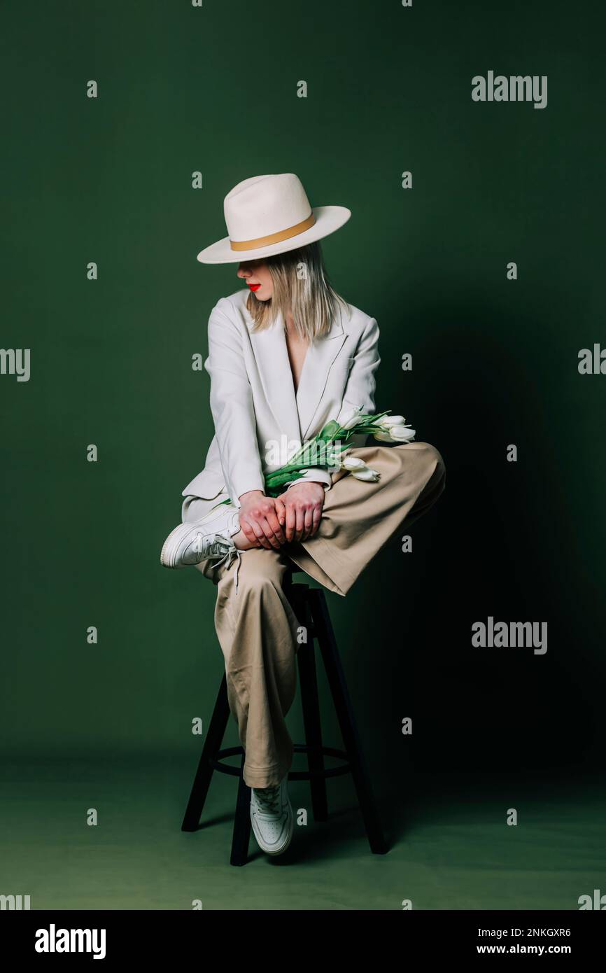 Fashionable woman with flowers wearing hat on stool in front of green ...