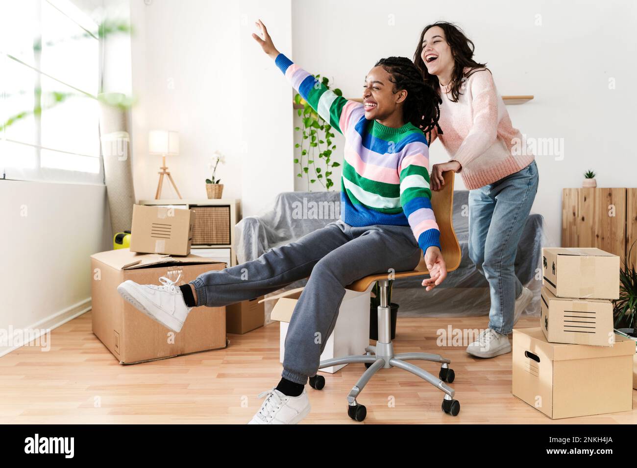 Carefree young woman pushing friend sitting in swivel chair at new home ...