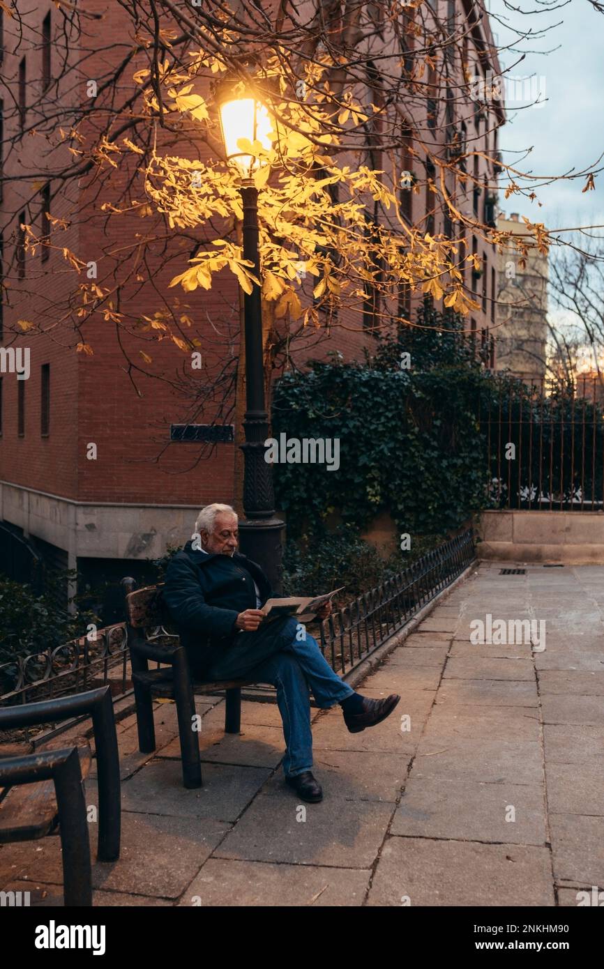 Senior man reading book sitting on chair near illuminated lamp post ...