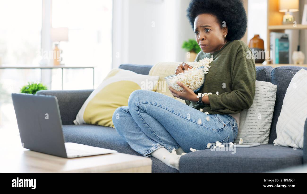 African woman watching a horror movie on a laptop and eating popcorn ...