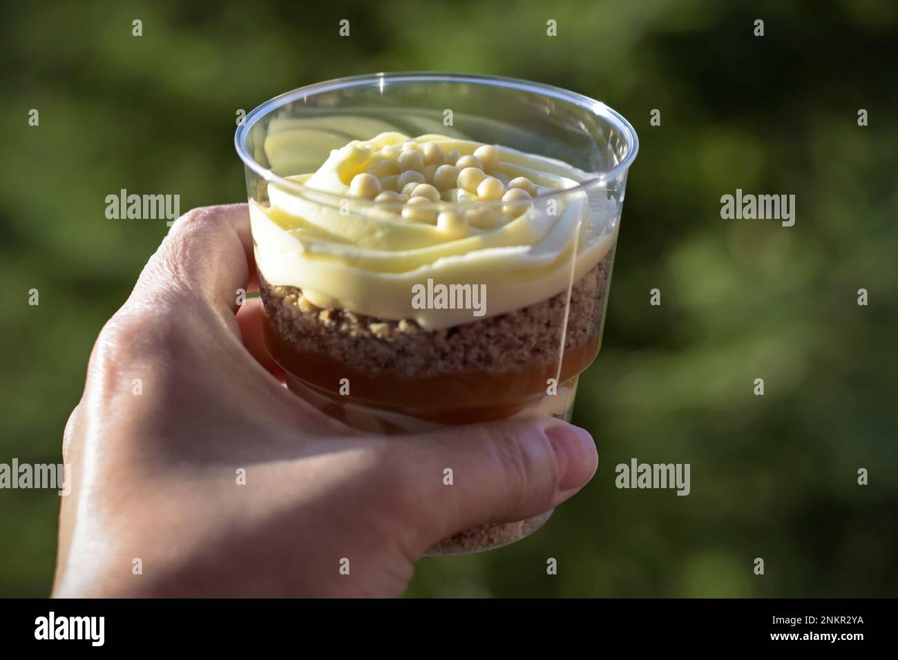 Sweet dessert in transparent glass in woman's hand against green background. Close-up. Trifle is ...