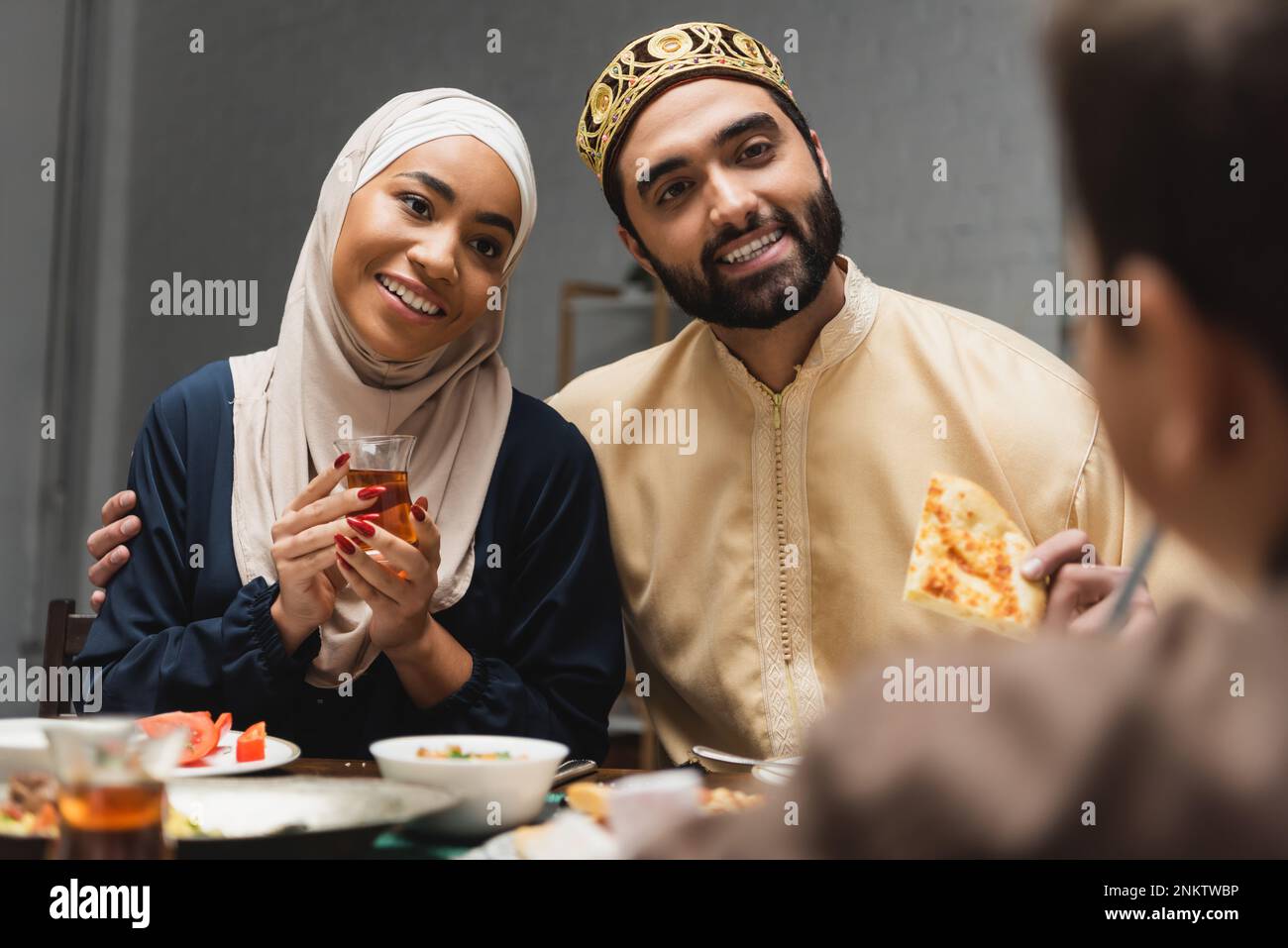 Smiling muslim parents holding food and tea near blurred child at home ...