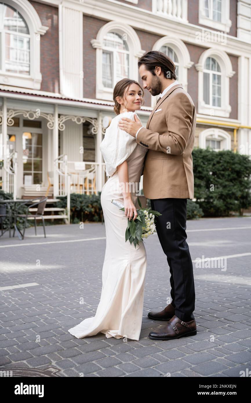 full length of bearded groom in suit hugging happy bride in white dress ...